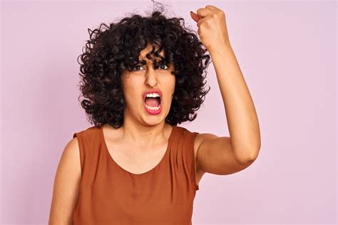 고립 된 분홍색 배경에 티셔츠를 입고 곱슬 머리를 가진 젊은 아랍 여성은 화가 난 좌절된 외침 미친 듯이 제기 된 손으로 고함