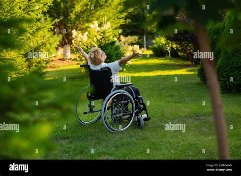 Rear View Of An Elderly Woman Spread Her Arms To The Sides While Sitting In A Wheelchair On A