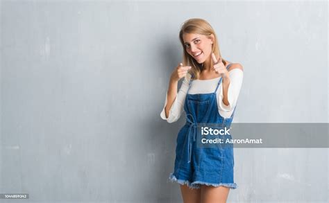 행복 하 고 재미 있는 얼굴로 카메라를 손가락을 가리키는 그런 지 회색 벽에 서 있는 아름 다운 젊은 여자 좋은 에너지와 느낌입니다 가리키기에 대한 스톡 사진 및 기타