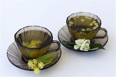 허브 핫 린든 차와 흰색 배경에 고립 된 꽃꽃이있는 재스민 차를 곁들인 두 개의 어두운 유리 컵 테이블에 녹색 잎검은 머그잔에 건강한 음료 복사 공간을 복사합니다 0명에