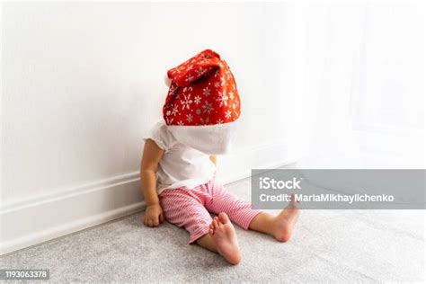빨간 산타 클로스 모자를 쓴 아기는 흰색 벽 근처의 회색 카펫에 앉아 크리스마스를 기다립니다 새해 컨셉입니다 재미 아이 피곤