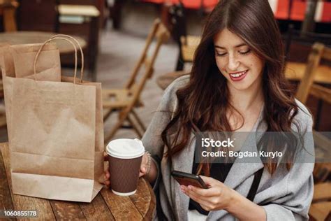 아름 다운 머리카락과 미소 전화 검색 커피 컵과 거리에 테라스에서 나무 테이블에 가방 유행 Hipster 소녀 화려한 행복 한 젊은 여자 웃 고 즐기는 시간 갈색에 대한
