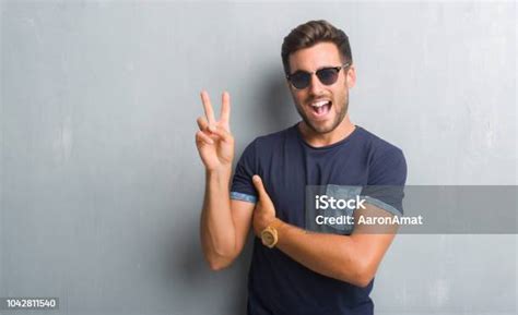 선글라스 승리 표시를 하 고 카메라에 윙크 하는 행복 한 얼굴로 미소를 입고 회색 그런 지 벽에 잘생긴 젊은 남자 번호 2입니다 긍정적인 감정 표현에 대한 스톡 사진 및