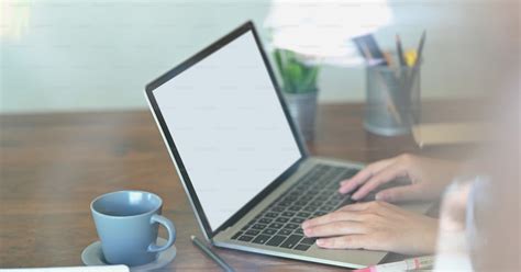 화분에 심은 식물 연필꽂이 커피 컵으로 둘러싸인 나무 작업 책상에 올려놓은 흰색 빈 화면 컴퓨터 노트북에 타이핑하는 아름다운 여성의 자른 이미지 사진