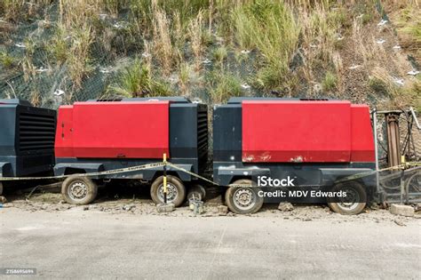 길가에 있는 대형 빨간색 산업용 휴대용 디젤 엔진 발전기가 주의 테이프와 금속 울타리로 줄지어 서 있습니다 도로 및 Riprap 건설 현장의 스크류 공기 압축기 연료 및