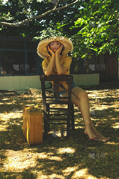 一个乡村妇女坐在木椅上，戴着草帽，穿着黄色衬衫和牛仔短裤。