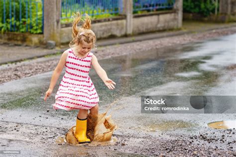 노란 장화를 신고 웅덩이를 걷는 행복한 미취학 소녀 화려한 옷을 입은 귀여운 아이가 웅덩이에 뛰어들고 물을 튀기고 야외 활동을 한다 건강한 생활방식에 대한 스톡 사진 및