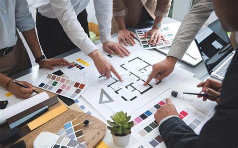 A Group Of Designers Collaborate Around A Table Reviewing A Floor Plan And Color Swatches For