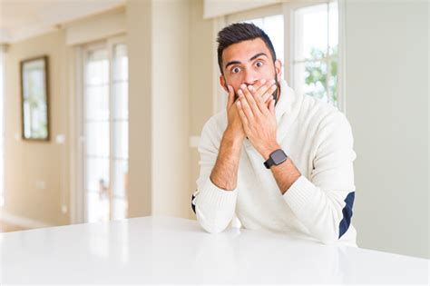 집에서 캐주얼 흰색 스웨터를 입고 잘생긴 히스패닉 남자는 실수로 손으로 입을 덮고 충격을 받았다 비밀 개념입니다 가리기에 대한 스톡 사진 및 기타 이미지 Istock