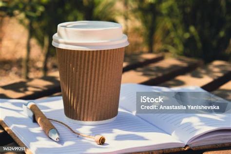 나무 벤치에 빈 종이 노트북이 있는 크래프트 종이에 커피 또는 차가 있는 에코 재활용 종이컵 야외에서 일하는 연구의 개념 커피를 가져 가라 텍스트 공간을 복사합니다 일회용