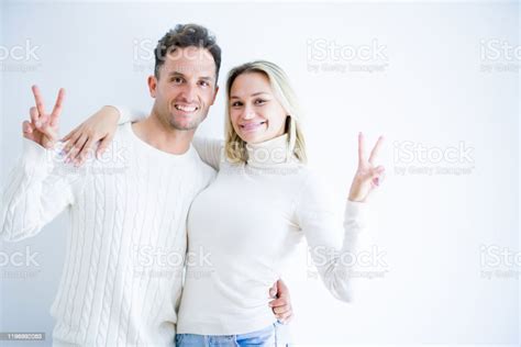 고립 된 흰색 배경 위에 서있는 캐주얼 한 티셔츠를 입은 젊은 아름다운 부부는 승리의 표지판을하고있는 카메라에서 행복한 얼굴 윙크와 미소 2번 2명에 대한 스톡 사진 및