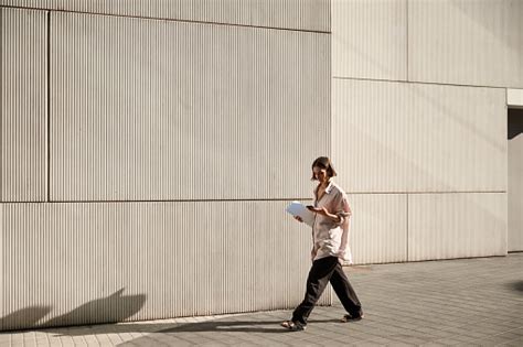 미니멀한 도시 환경에서 휴대폰을 사용하여 걷는 전문 사업가로서 현대적인 비즈니스 활동과 도시 건축의 조화를 보여줍니다 Brand Name Smart Phone에 대한 스톡