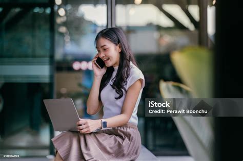 노트북을 사용하고 스마트 폰으로 통화하면서 원격으로 작업하고 도시 환경에서 야외 벤치에 앉아 현대적인 라이프 스타일을 즐기는 쾌활한 젊은 아시아 여성 30 34세에 대한