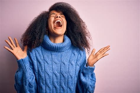 젊은 아름 다운 아프리카 계 미국인 여자 Afro 머리 를 입고 겨울 스웨터 를 입고 분홍색 배경에 미친 축 하 와 성공을 위해 미친 팔을 올리고 눈을 감고 흥분 비명을