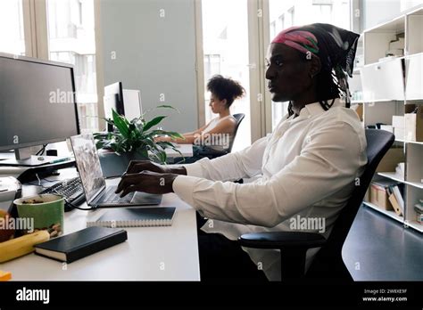 Male Computer Programmer Using Laptop While Sitting On Chair With Colleague At Office Stock