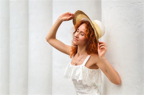 닫아 잘 손질 된 빨간 머리 곱슬 소녀는 흰색 기둥 근처에서 포즈를 취합니다 그녀는 꿈꾸며 눈을 감고 모자를 들고 있다 공간을 복사합니다 가냘픈에 대한 스톡 사진 및 기타