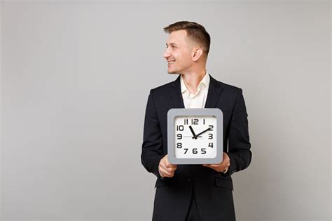 고전적인 검은 양복과 흰색 셔츠에 젊은 비즈니스 남자는 스튜디오에서 회색 벽 배경에 고립 된 사각형 시계를 들고 옆으로 찾고