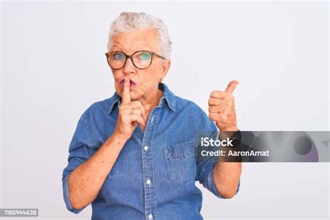 데님 셔츠와 안경을 쓰고 고립된 흰색 배경 위에 회색 머리의 선임 여성이 손을 가리키는 입술에 손가락으로 조용히 하라고 요구합니다 침묵과 비밀 개념 가리키기에 대한 스톡