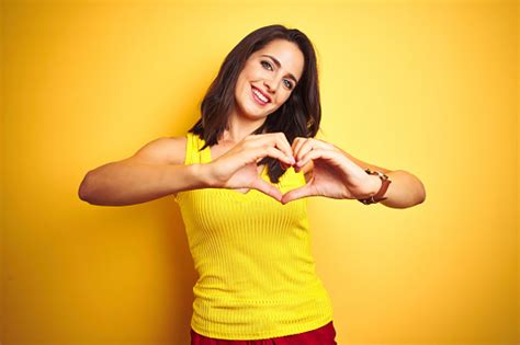 노란색 고립 된 배경 위에 서있는 티셔츠를 입은 젊은 아름다운 여성은 손으로 심장 기호 모양을하고 사랑에 미소 로맨틱 한 개념 만들기에 대한 스톡 사진 및 기타 이미지