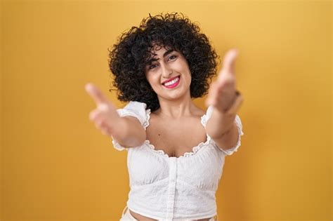 곱슬 머리가 노란 배경 위에 서있는 젊은 갈색 머리 여자가 포옹을 위해 두 팔을 벌려 웃고있는 카메라를 바라 봅니다 행복을 포용하는 쾌활한 표현 손에 대한 스톡 사진 및