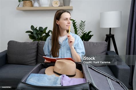 아늑한 소파에 앉아 꿈꾸는 젊은 여성은 메모를 하고 목록을 확인하고 수하물에 옷을 포장하거나 준비하고 여행 여행을 위한 여행용 케이스를 준비합니다 여행 개념의 배낭 여행