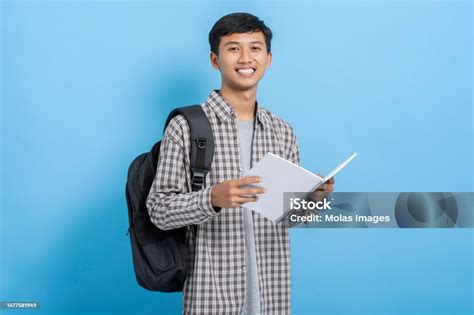 플란넬 셔츠를 입은 젊은 아시아 대학생이 배낭을 들고 파란색 배경 위에 고립된 책을 읽고 있다 18 19세에 대한 스톡 사진 및