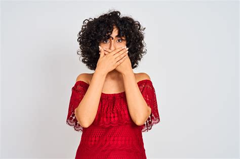 고립 된 흰색 배경 위에 서있는 빨간 드레스를 입은 곱슬 머리를 가진 젊은 아랍 여성은 실수로 손으로 입을 덮는 충격을