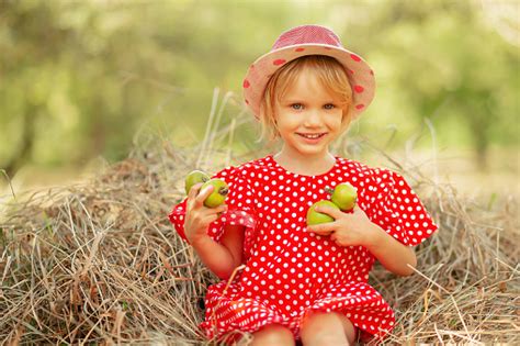 물방울 무늬 빨간 드레스와 밀짚 모자에 있는 리틀 소녀가 건초 더미에 앉아 사과를 들고 있다 금발 머리와 푸른 눈 재미 아이