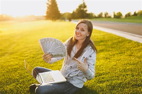 달러 현금 돈을 여성 지주 번들 여자 잔디에 앉아 푸른 잔디 햇빛 잔디 야외에 도시 공원에서 노트북 Pc 컴퓨터에서 작업 모바일 사무실입니다 프리랜서 비즈니스 개념입니다