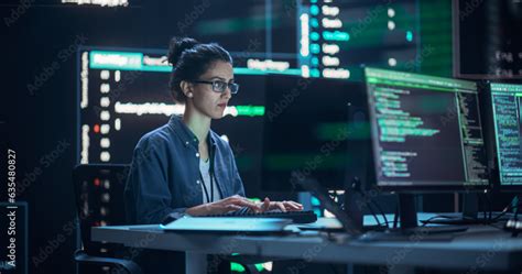 Female Programmer Working In Monitoring Control Room Surrounded By Big Screens Displaying Lines