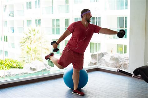 분홍색 티셔츠와 분홍색 안경을 쓴 장난스러운 뚱뚱한 남자는 체육관에서 아령과 핏 볼로 피트니스에 종사하고 있습니다 체육관에서 물을 마시는 남자 헤드폰으로 음악을 듣는 남자