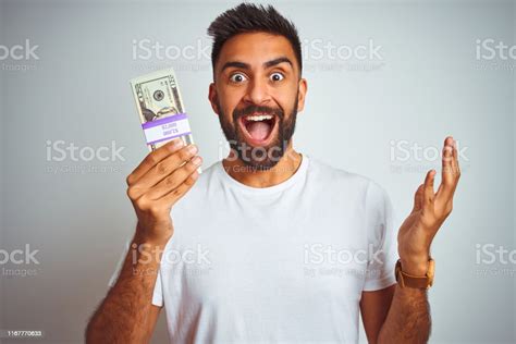 고립 된 흰색 배경 위에 서있는 달러를 들고있는 젊은 인도 남자는 매우 행복하고 흥분 큰 미소와 함께 비명을 지르는 승리를 축하하는 승자 표정 금융에 대한 스톡 사진 및