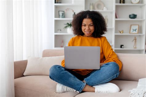 바쁘고 쾌활한 예쁜 밀레니엄 세대 아프리카 계 미국인 여성이 미니멀리스트 거실 인테리어에서 컴퓨터에서 타이핑하고 있습니다 공부 프리랜서 집에서 원격으로 일하고 비즈니스