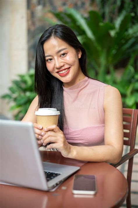 A Beautiful Asian Woman Sitting At An Outdoor Table With Her Laptop Smiling At The Camera Stock