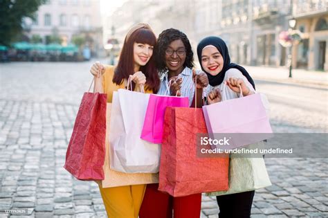 세련된 패션 옷을 입은 백인 아프리카 이슬람교도 등 아름다운 세 명의 다민족 소녀들이 오래된 유럽 도시에서 야외에서 서서 미소짓고 있는 쇼핑백을 들고 있다 3 명에 대한