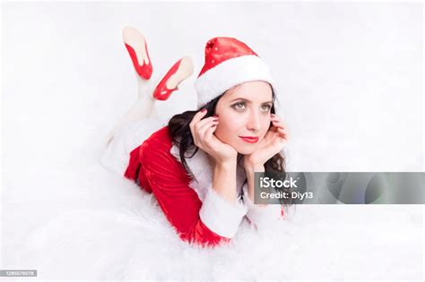 산타 모자를 쓴 젊은 여성이 침대에 누워 서서 꿈꾸며 창밖으로 바라보고 있습니다 크리스마스를 기다리는 가정 환경 흰색 모피 담요에 누워 산타 클로스의 옷을 입은 소녀 흰색