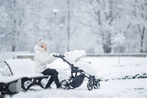 눈보라가 자자한 후 하얀 겨울날 마을 눈 덮인 공원에서 벤치에 앉아 있는 젊은 성인 여성 어머니 옆에 유모차 측면 보기입니다 자연의 평화로운 분위기 겨울에 대한 스톡 사진