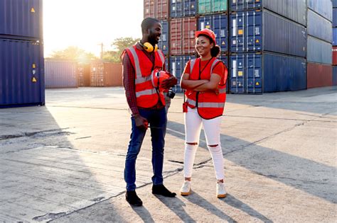 두 명의 행복한 미소 산업 아프리카 엔지니어 남자와 여자는 안전 조끼와 헬멧을 착용하고 황혼 일몰 시간에 물류 선적화물 컨테이너 야드에서 함께 일하는 노동자 팀입니다 태양