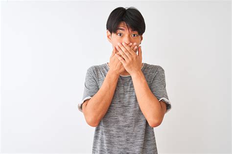 고립 된 흰색 배경 위에 서있는 캐주얼 한 티셔츠를 입은 젊은 중국 남자는 실수로 손으로 입을 덮는 충격을 받았습니다 비밀 개념입니다 T 셔츠에 대한 스톡 사진 및 기타