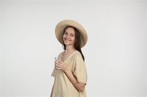 긴 머리를 한 젊은 여성이 한 손에 유리잔을 들고 미소를 짓고 있다 밝은 색의 의상에 챙이 넓은 밀짚모자를 쓴 그녀는 은은한 조명이 켜진 스튜디오에 서 있다 30 34세에