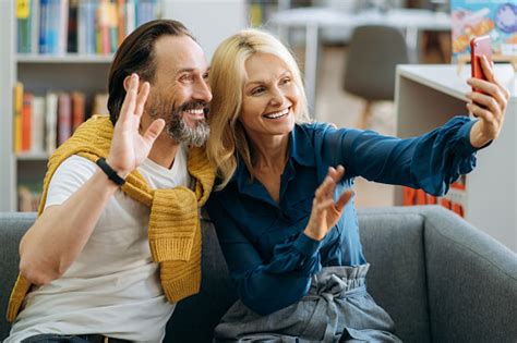 성숙한 친구들은 소파에 함께 앉아있다 수염과 아름다운 금발의 여자와 세련된 백인 자신감 남자는 소파에 앉아 스마트 폰을 사용하여 비디오 통신에 채팅 미소 자신감에 대한 스톡