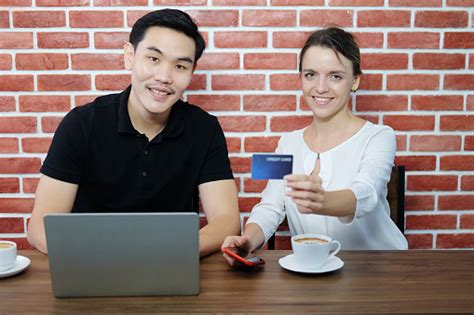 미소 짓는 젊은 비즈니스 부부는 아시아 남자와 백인 여성이 온라인 쇼핑을위한 신용 카드를 들고 커피 숍의 나무 테이블에 노트북으로 소셜 미디어에서 지불하는 것을 좋아합니다