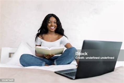 흰색 티셔츠와 청바지를 입은 젊은 아프리카 계 미국인 소녀는 침실에서 노트북 Pc와 메모장이있는 침대에 앉아 집에서 공부하거나 일하고 집에서 시간을 보내고 있습니다 온라인