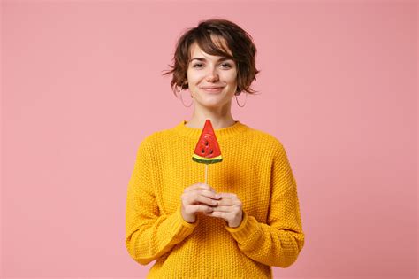 스튜디오에서 파스텔 핑크 벽 배경에 고립 포즈 노란색 스웨터에 젊은 갈색 머리 여자 소녀 미소 사람들은 진지한 감정 라이프 스타일 개념 복사 공간을 모의합니다 수박 롤리팝을