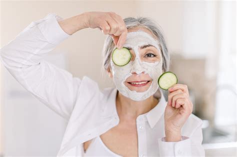 얼굴 진흙 마스크와 오이 슬라이스와 함께 미소 예쁜 선임 회색 머리 여자의 아름다움 초상화를 닫고 밝은 집 인테리어 배경에 포즈 스킨 코스메틱 과 스파 컨셉 가정의 방에