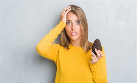 머리에 손으로 압박 하는 아보카도 먹는 그런 지 회색 벽에 아름 다운 젊은 여자는 수치심과 깜짝 얼굴로 충격 분노와 좌절 두려워 하 고 실수에 대 한 화가 감정에 대한 스톡