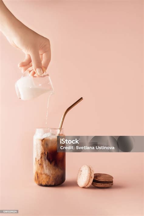 젊은 여성의 손이 황금 빨대와 키가 큰 유리에 아이스 커피 한 잔에 우유를 붓는 커피와 마카롱 초콜릿 파스텔 배경에 바닐라 디자인 빈티지 스타일의 음식 컨셉 각얼음에 대한