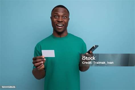 검은 피부를 가진 30세의 잘생긴 미국 남성은 스마트폰과 복사 공간이 있는 배경에서 쇼핑을 위한 플라스틱 카드를 들고 기본 티셔츠를 입고 있다 인사장에 대한 스톡 사진 및