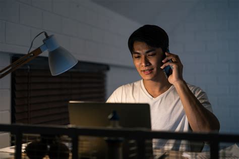 아시아 청년이 과로하고 밤에 휴대 전화로 통화합니다 직장에서 스마트폰 통화를 사용하는 매력적인 미남 사업가가 집에서 취침 시간 동안 테이블에서 온라인으로 의사 소통을 합니다