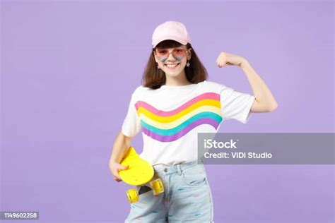 생생한 옷에 웃는 십 대 소녀 노란색 스케이트 보드를 들고 안경 팔 뚝을 보여주는 보라색 파스텔 배경에 고립 된 근육 사람들은 진심으로 감정 라이프 스타일 개념입니다 복사